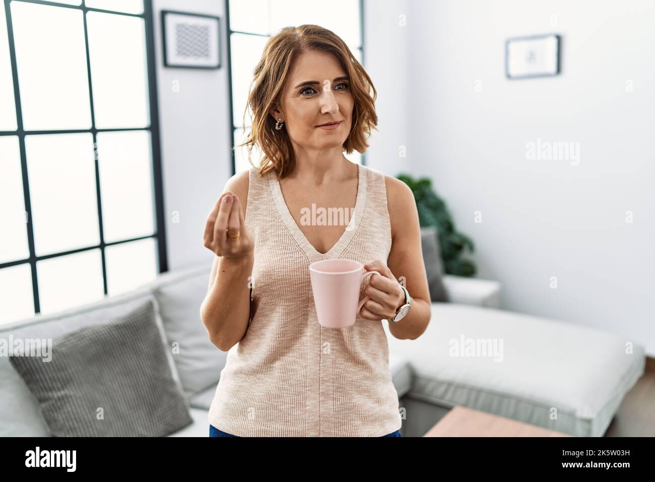 Middle age woman drinking a cup coffee at home doing money gesture with ...