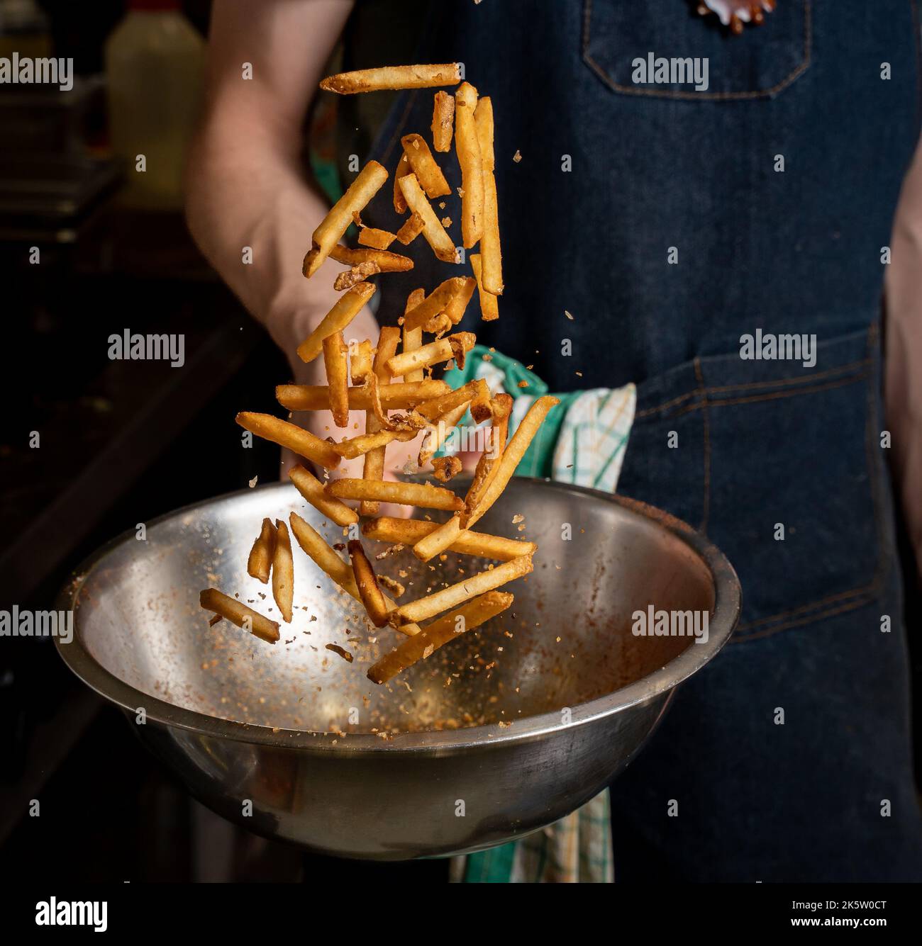 A person tossing freshly cooked fries with spices on a mixing bowl ...