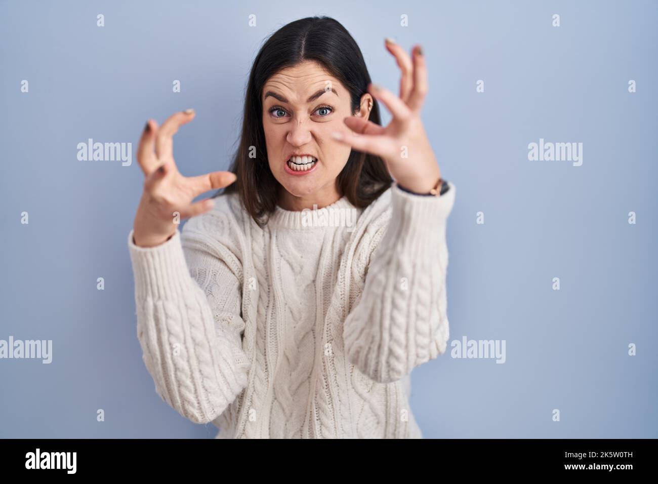 Young brunette woman standing over blue background shouting frustrated ...