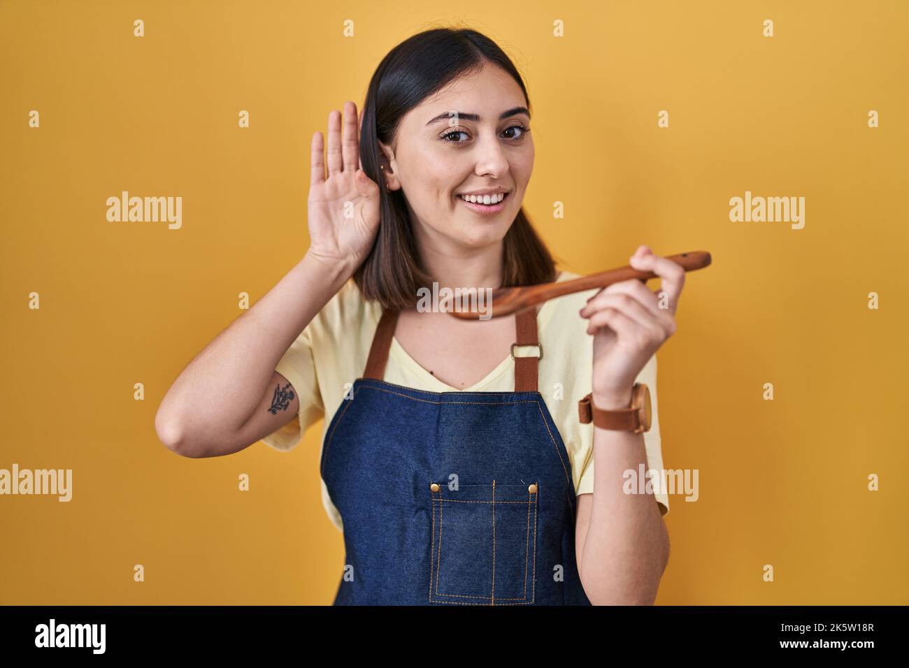 Hispanic girl eating healthy wooden spoon smiling with hand over ear ...