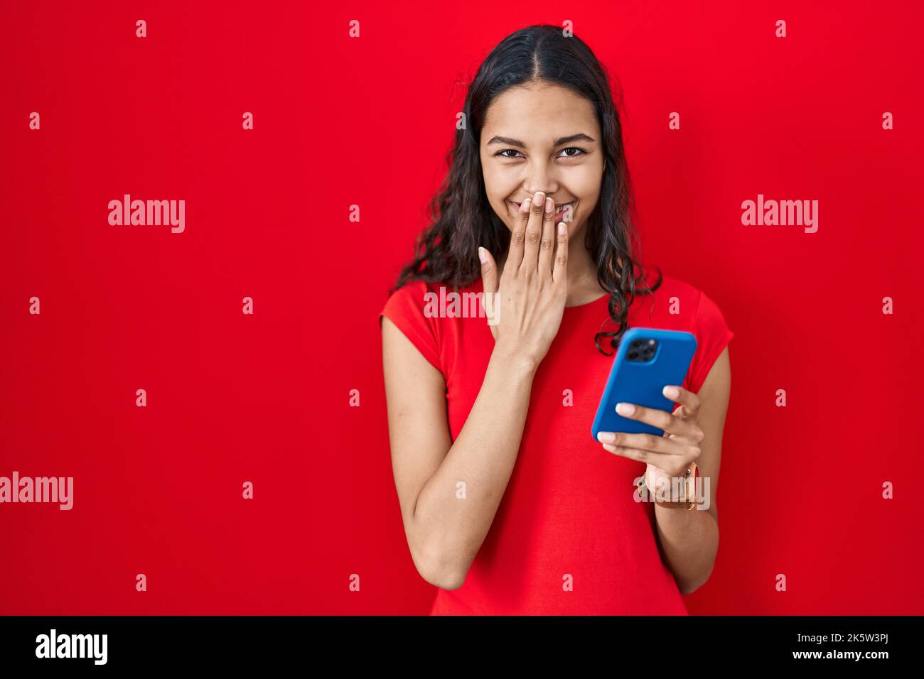 Young brazilian woman using smartphone over red background laughing and ...