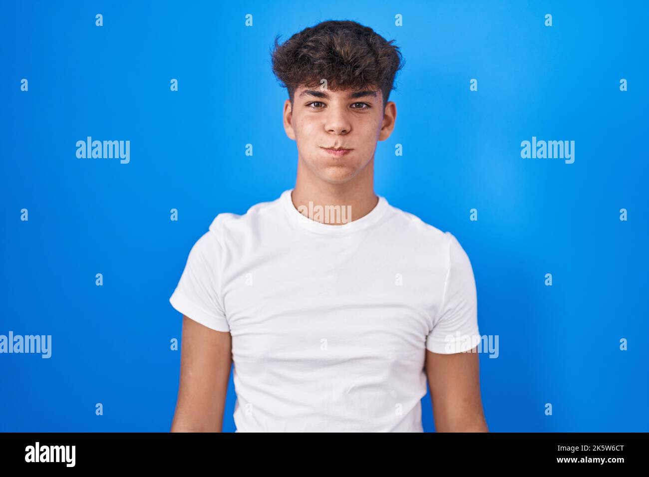 Hispanic teenager standing over blue background puffing cheeks with ...