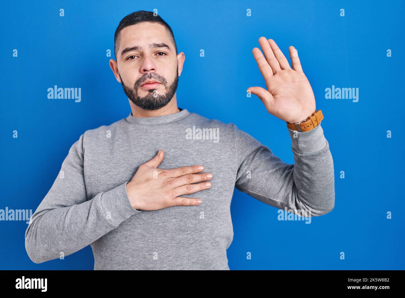 Hispanic man standing over blue background swearing with hand on chest ...