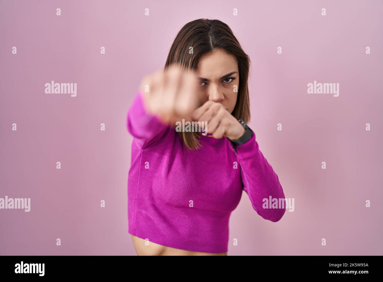 Hispanic woman standing over pink background punching fist to fight ...