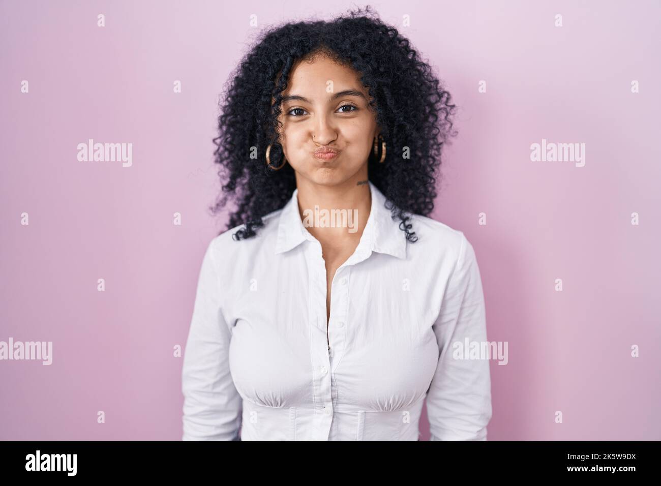 Hispanic woman with curly hair standing over pink background puffing ...