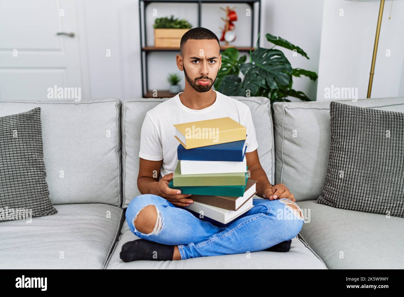 African american young man holding a pile of books sitting on the sofa ...