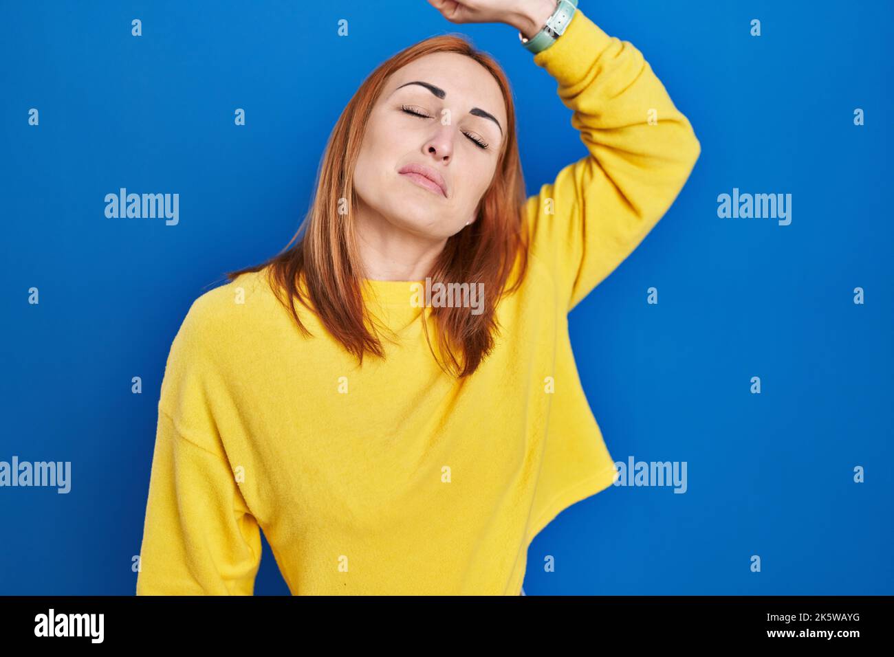 Young woman standing over blue background stretching back, tired and ...