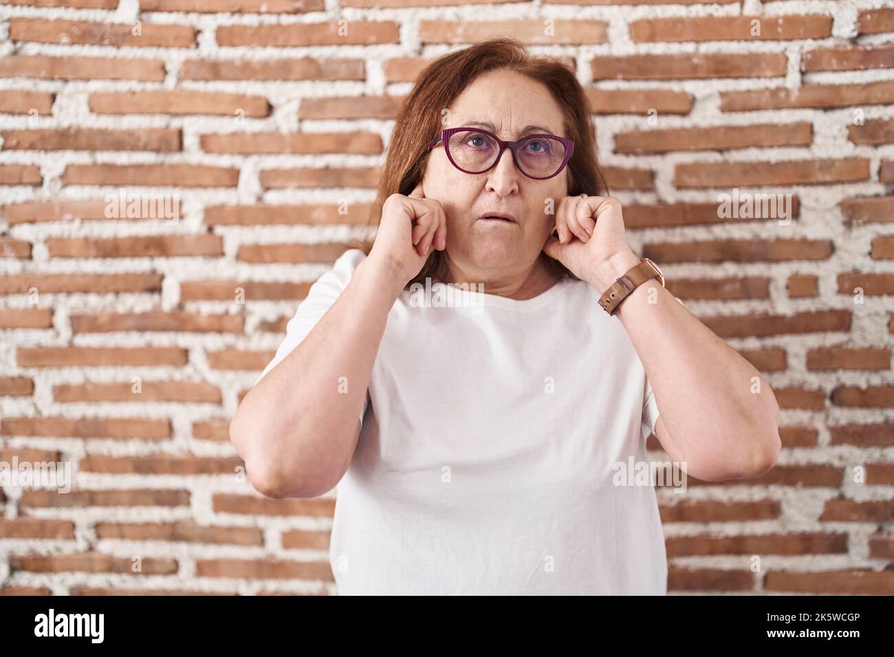 Senior woman with glasses standing over bricks wall covering ears with ...
