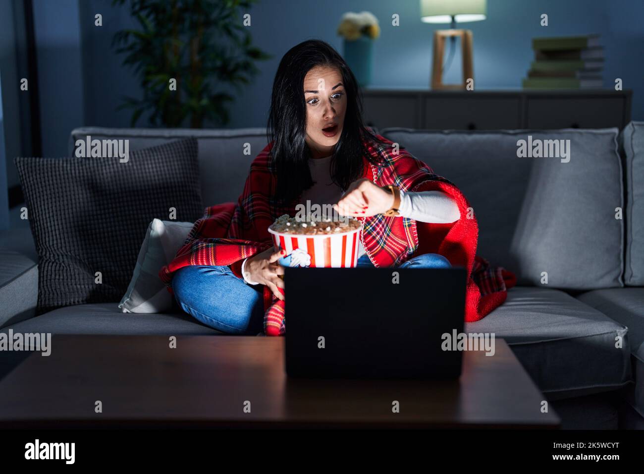 Hispanic woman eating popcorn watching a movie on the sofa looking at ...