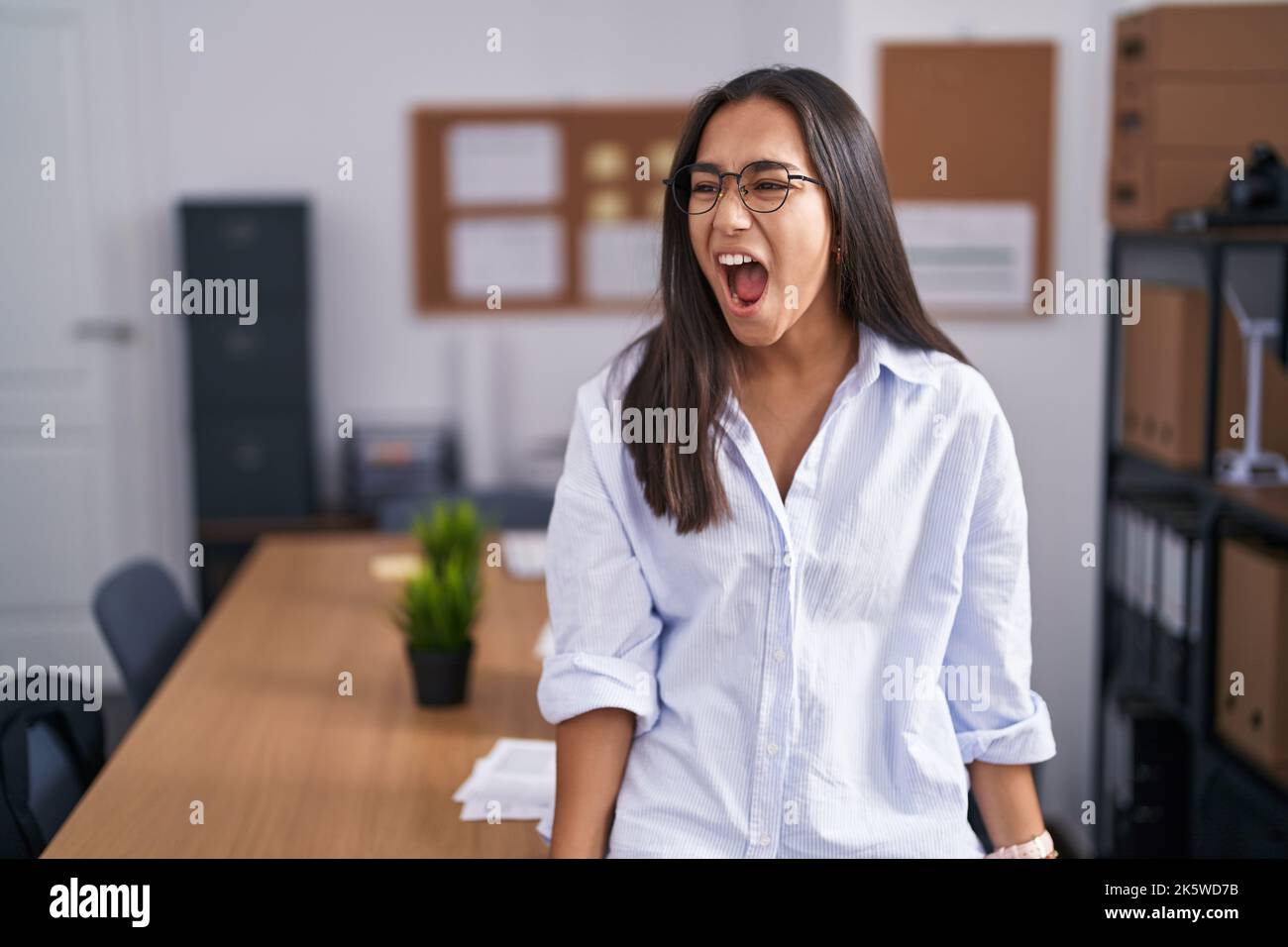 Young hispanic woman at the office angry and mad screaming frustrated ...