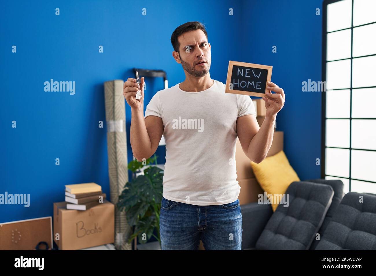 Young hispanic man with beard holding keys of new home in shock face ...