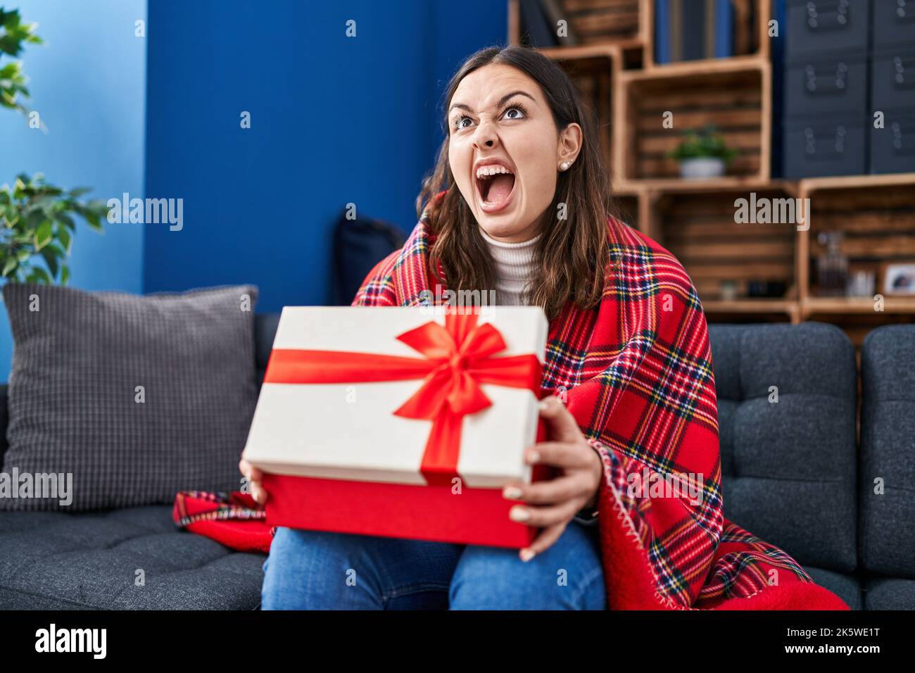 Young hispanic woman opening gift box angry and mad screaming ...