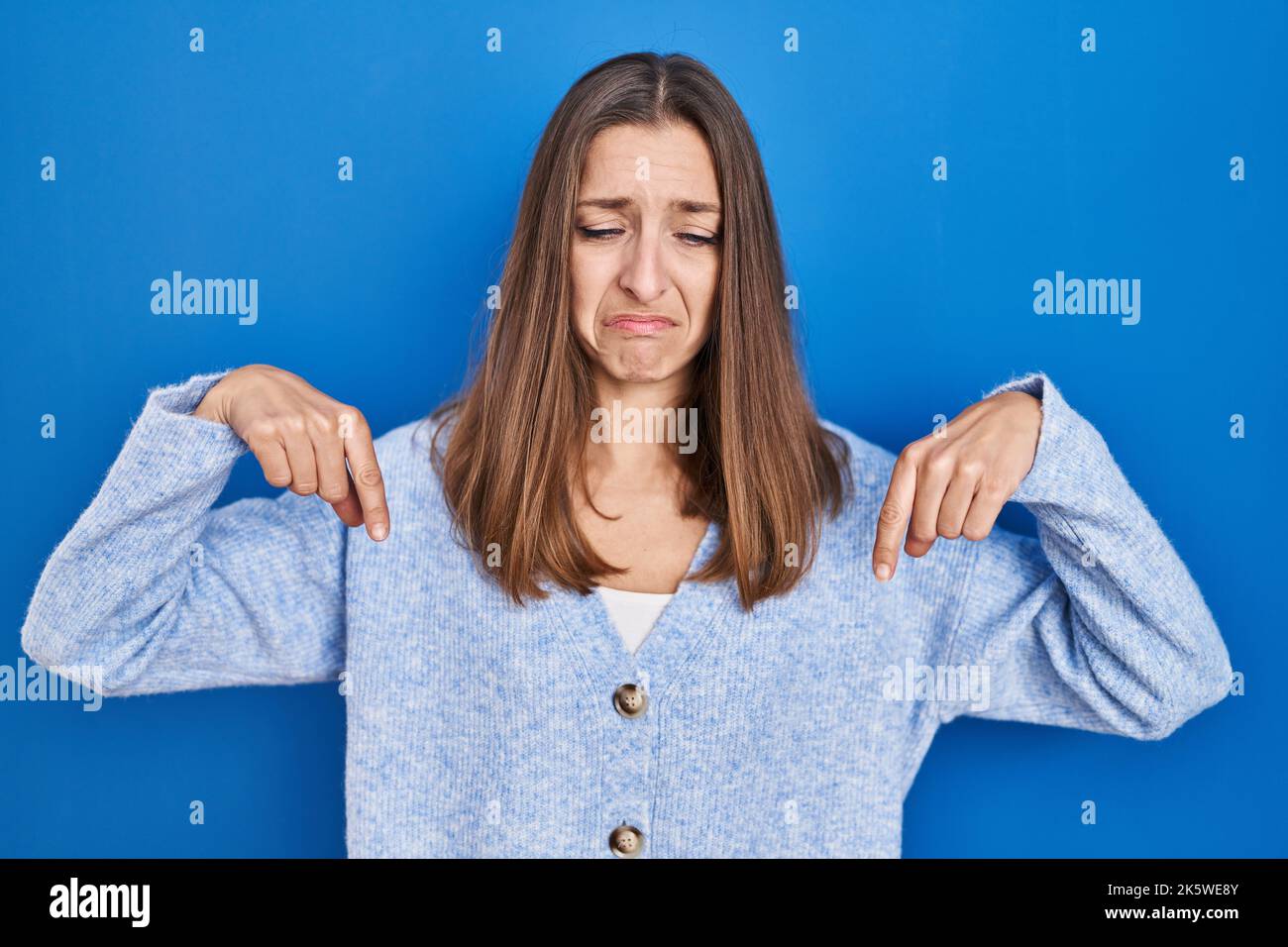 Young woman standing over blue background pointing down looking sad and ...