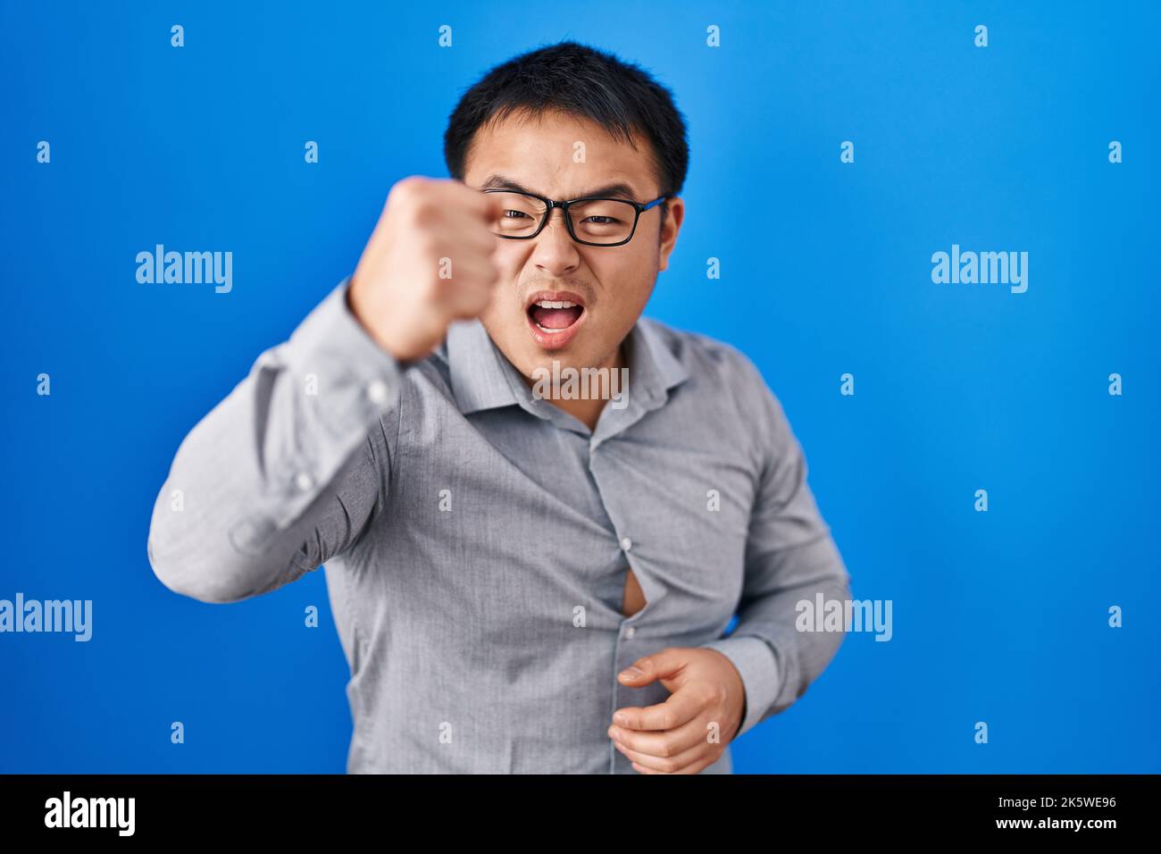 Young chinese man standing over blue background angry and mad raising ...
