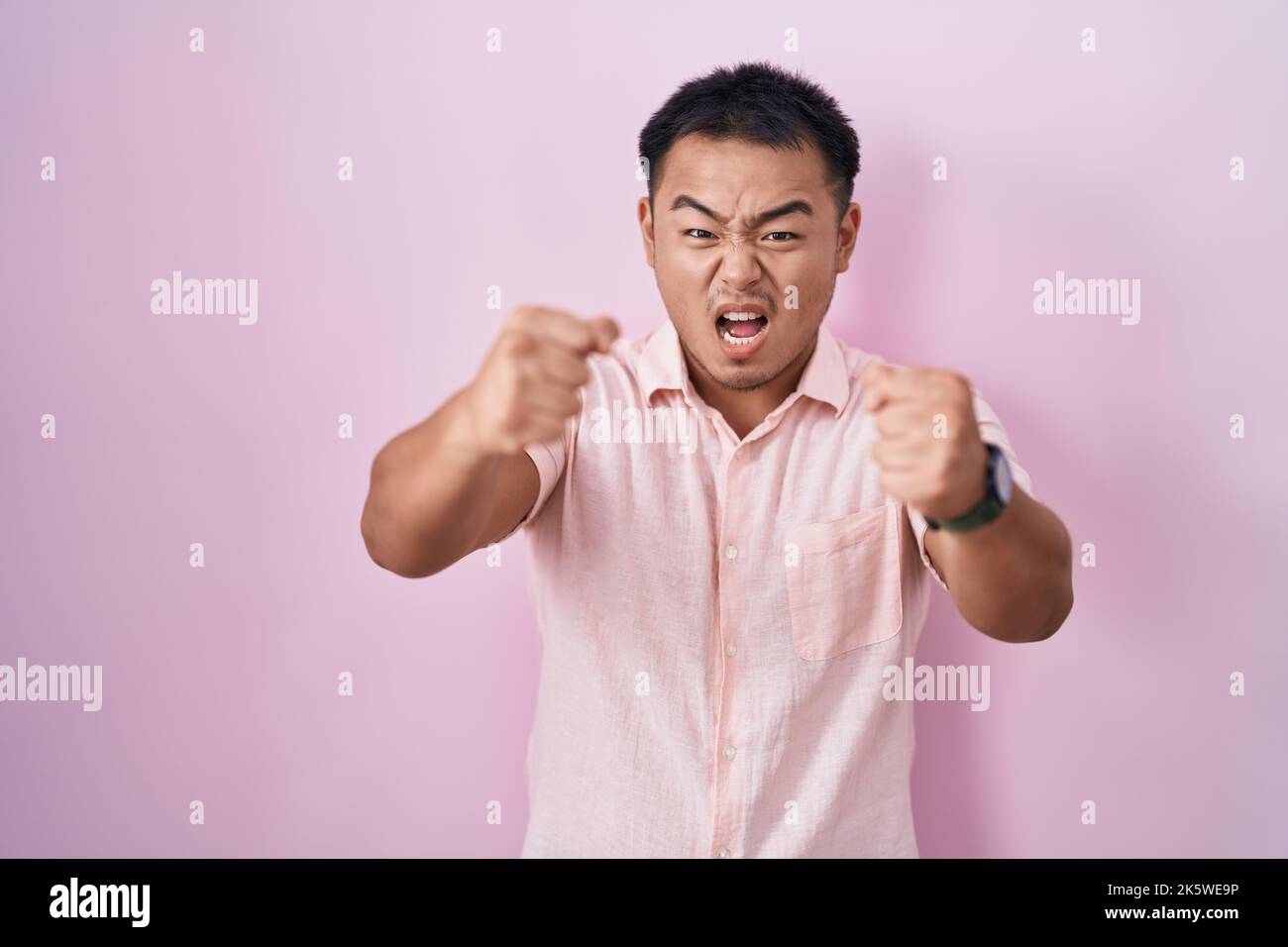 Chinese young man standing over pink background angry and mad raising ...