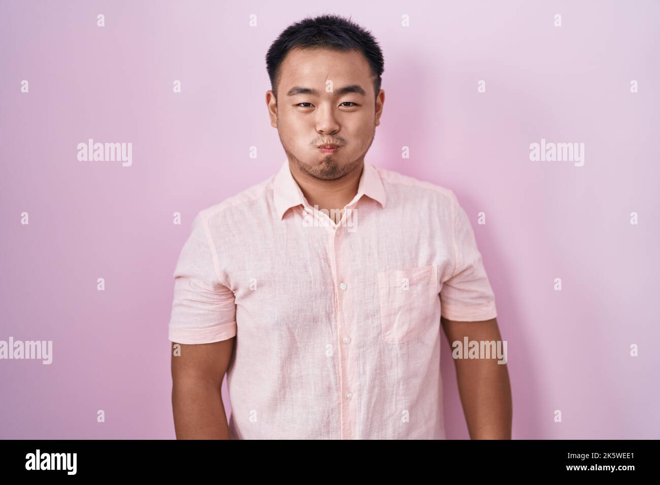 Chinese young man standing over pink background puffing cheeks with ...