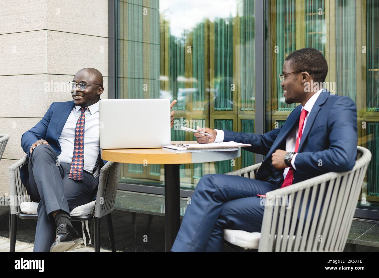 Two dark-skinned African American businessmen in suits and glasses with briefcases sit at a ...
