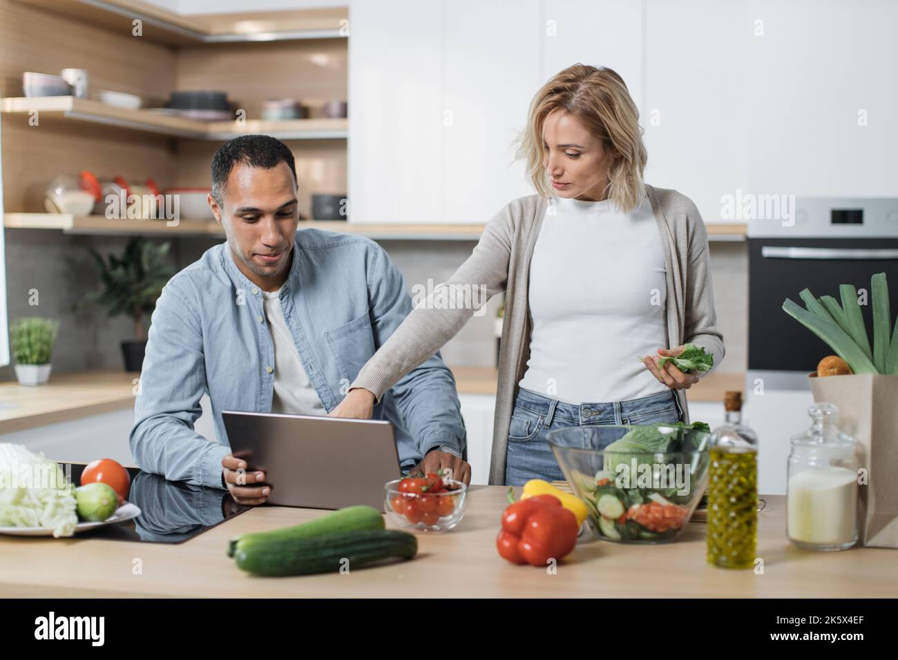 Happy married multinational couple using laptop while cooking healthy ...