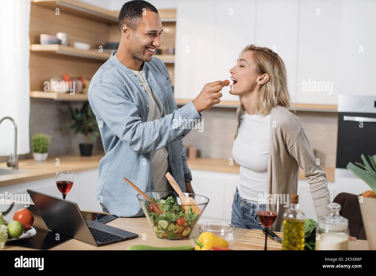 Cheerful married multinational couple using laptop while cooking ...