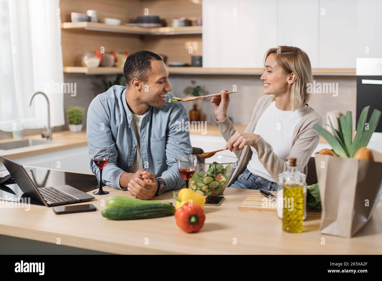 Cheerful married multinational couple using laptop while cooking ...