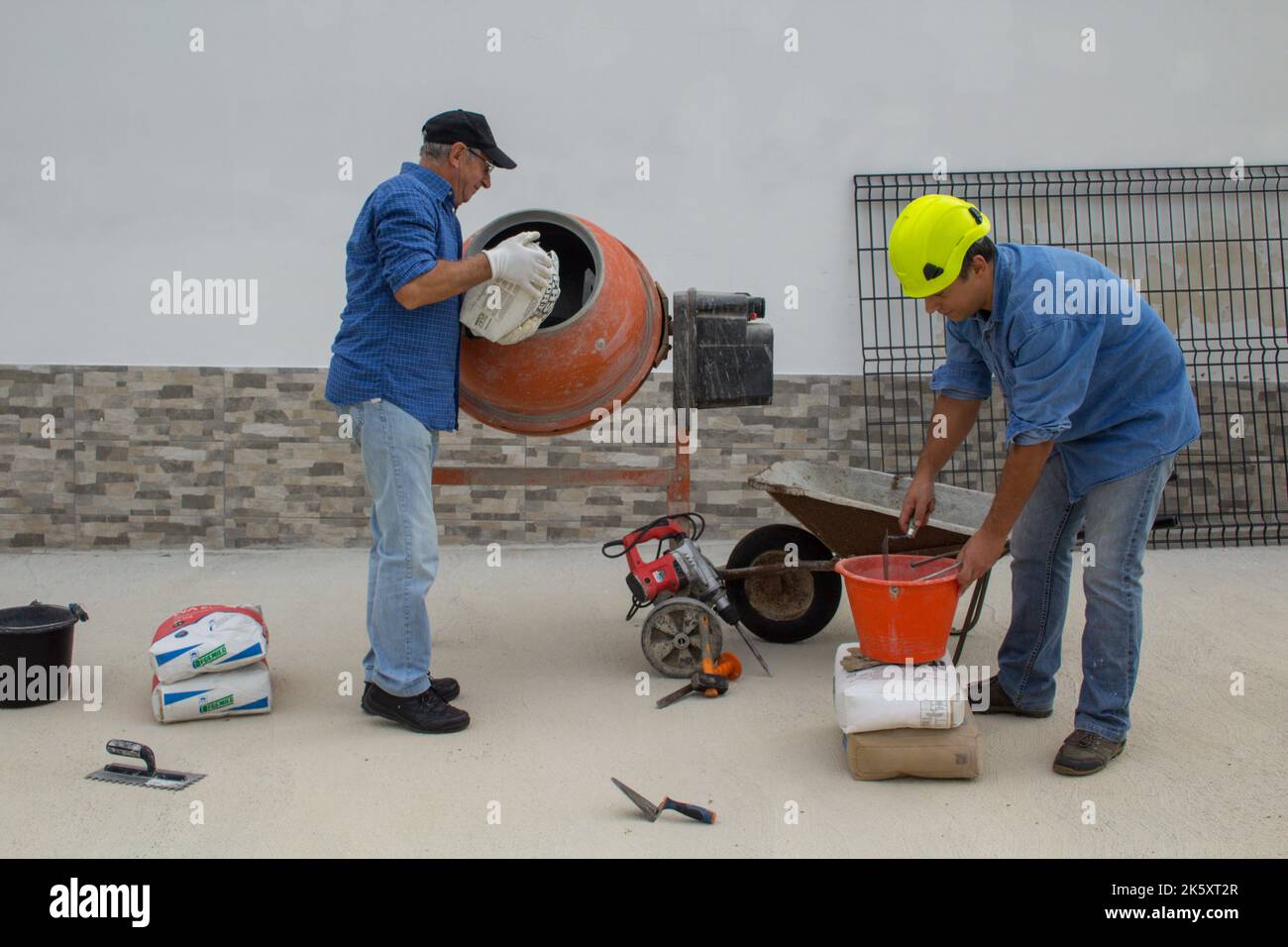 An elderly bricklayer and a young one are mixing concrete with a ...