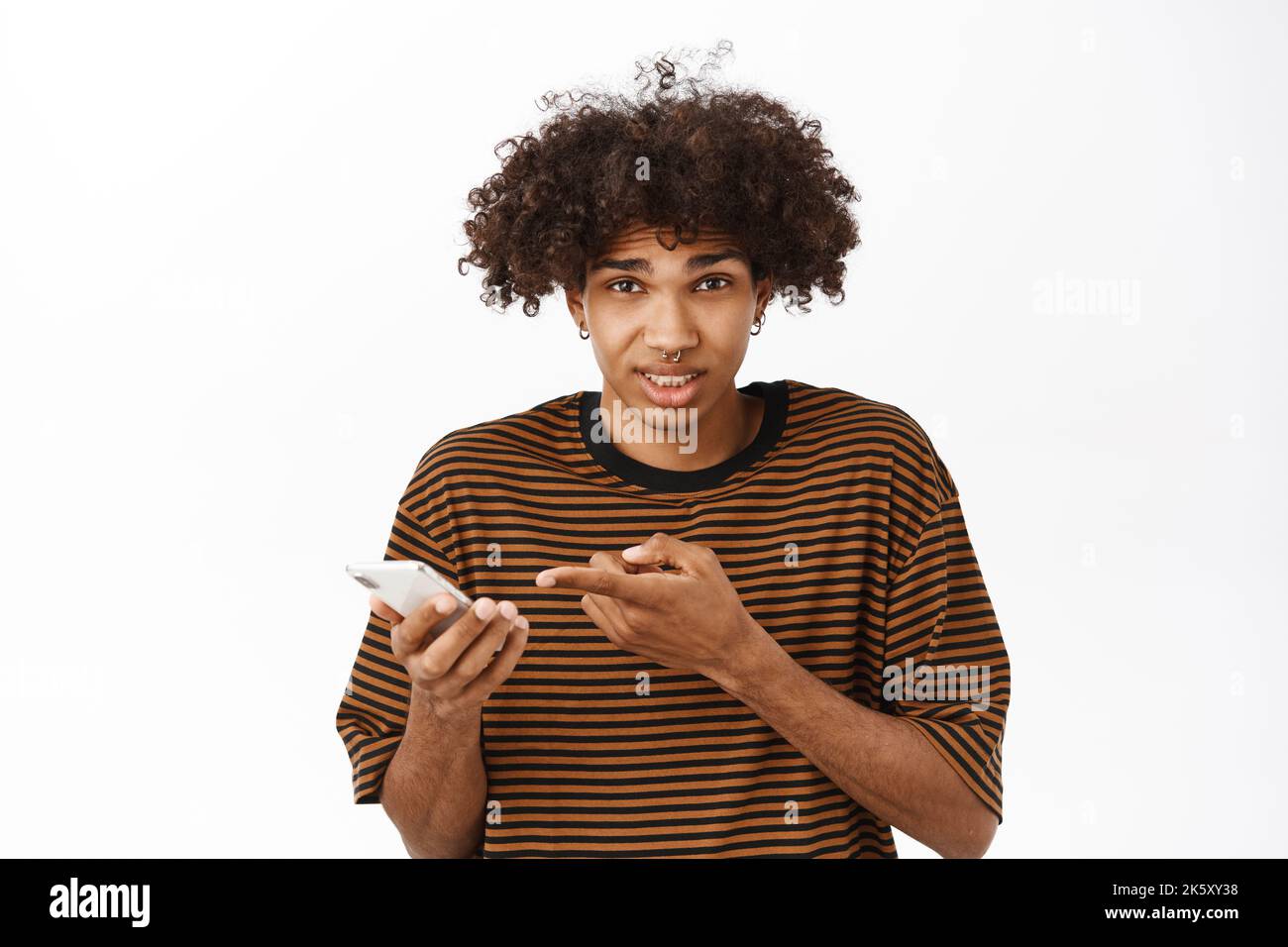 Portrait of young man holding phone, pointing at device with confused ...