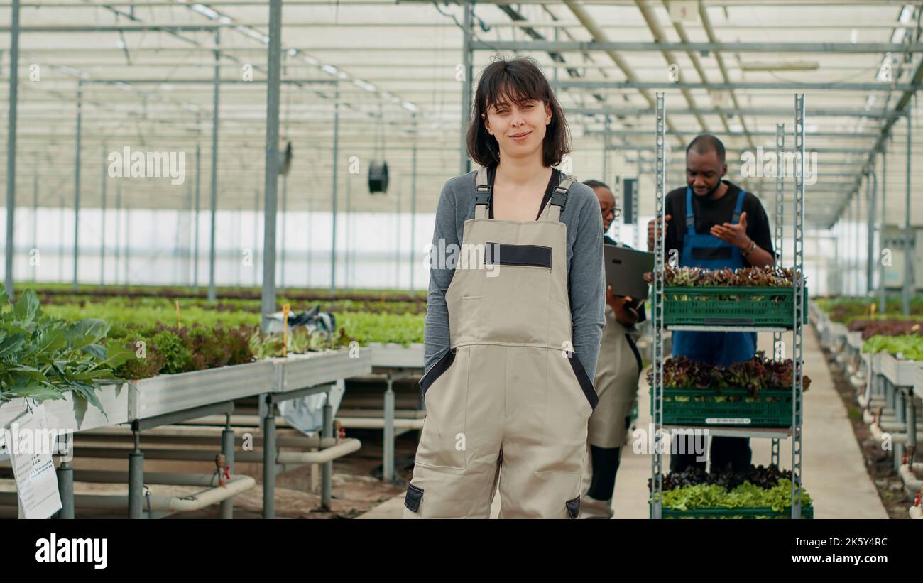 Portrait of confident woman posing with arms in pockets in greenhouse ...