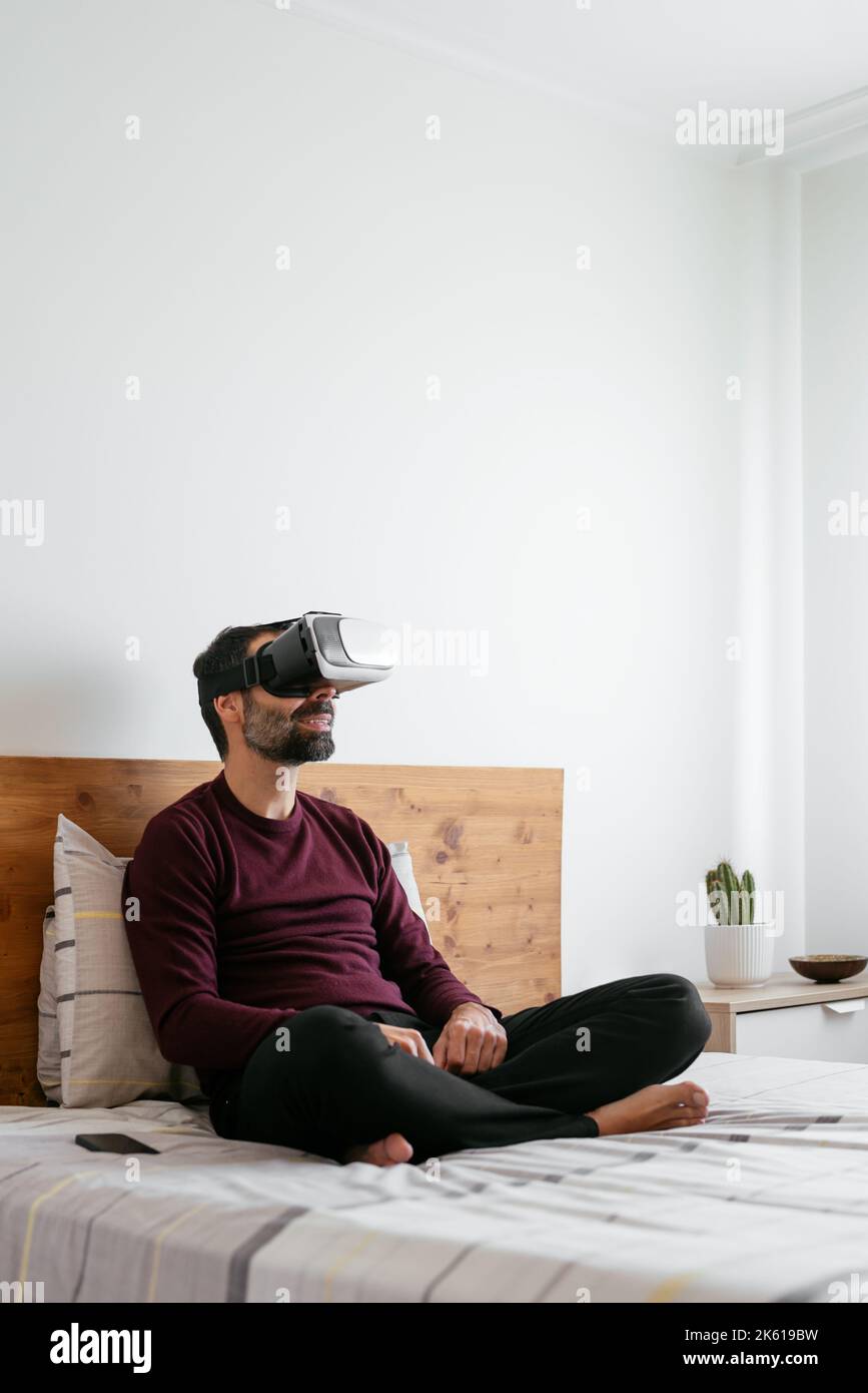 Man in VR headset sitting on bed with crossed legs in light cozy ...