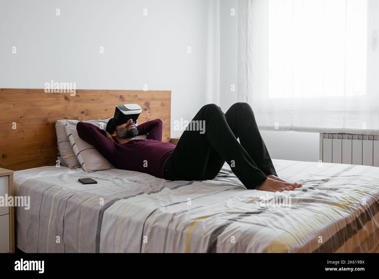Side view of guy with beard lying on bed and looking through VR goggles ...