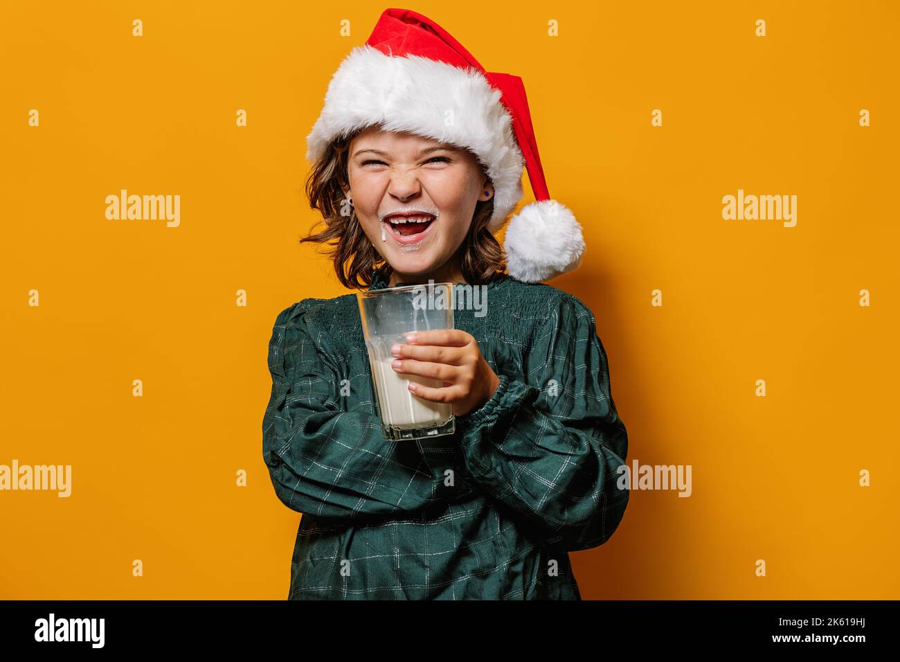 Cute cheerful girl in Santa hat sipping milk from glass and looking at ...