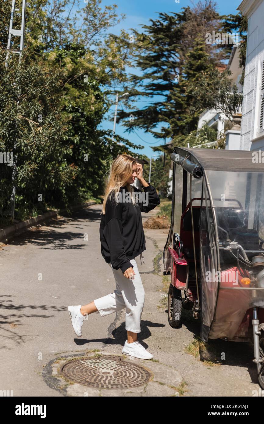 blonde woman in stylish outfit walking near three wheeled moped with ...