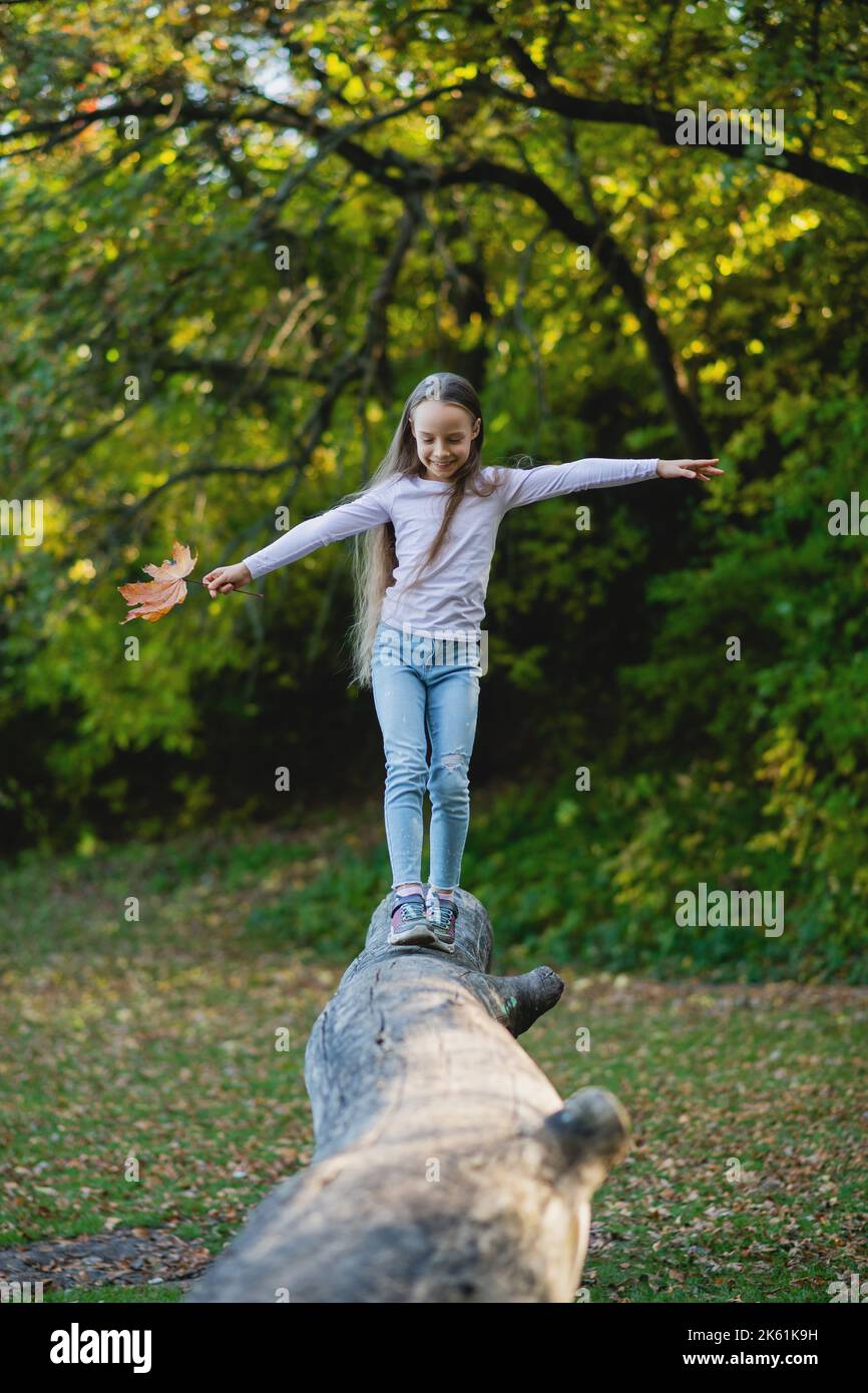 Cute girl balances on a fallen log with a leaf in her hand in the park ...
