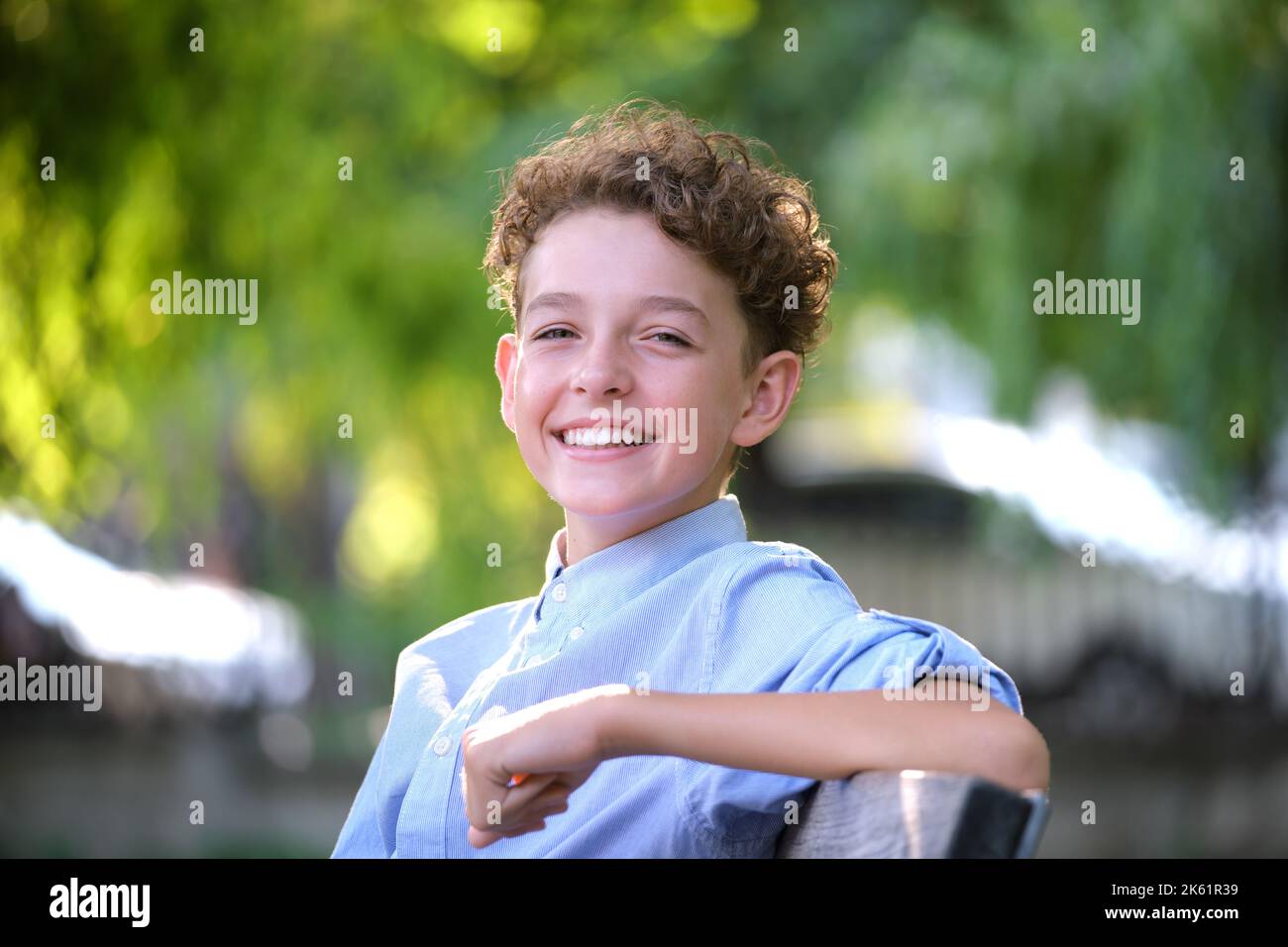 Young happy child boy relaxing sitting on bench in summer park. Positive kid enjoying summertime ...