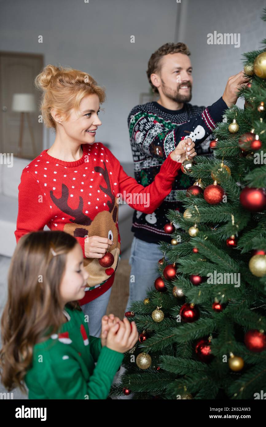 Positive woman in sweater decorating christmas tree with family at home ...