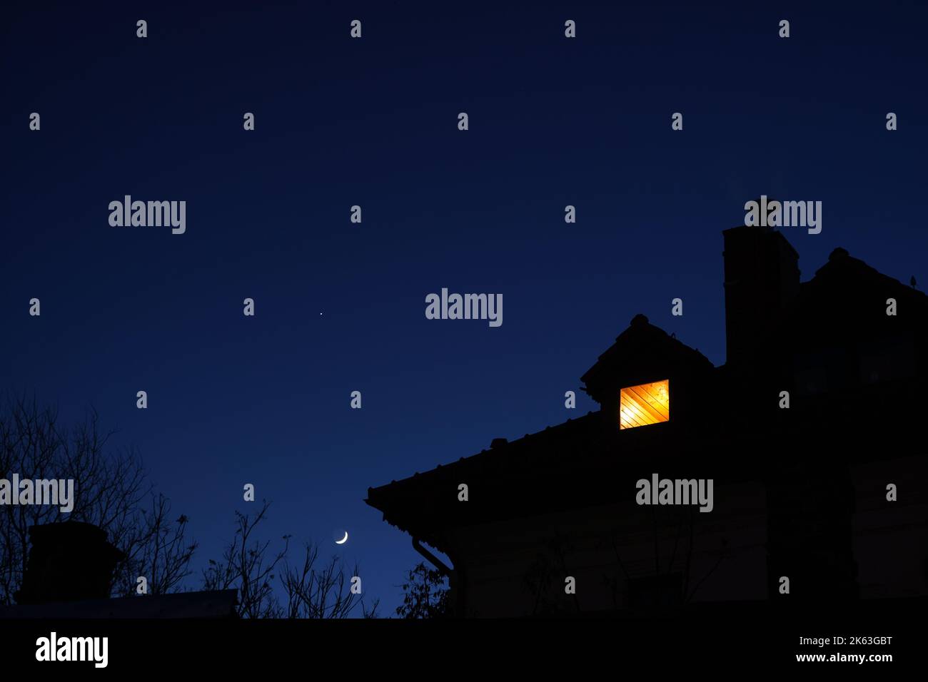 Silhouette of house roof with chimney and lit attic small window at ...