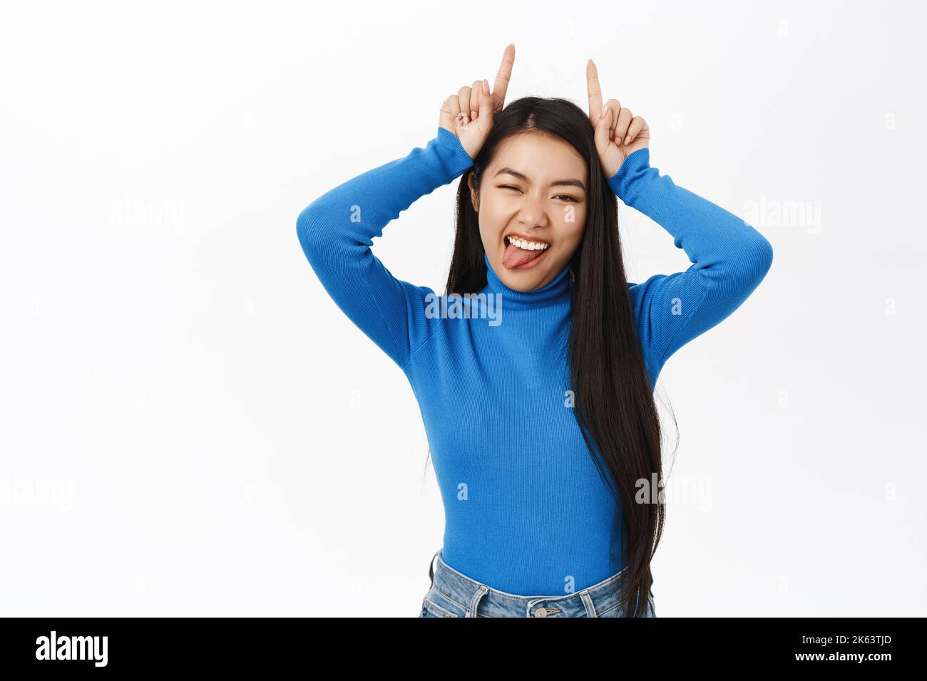 Enthusiastic asian woman shows devil horns, fingers on head, smiles ...