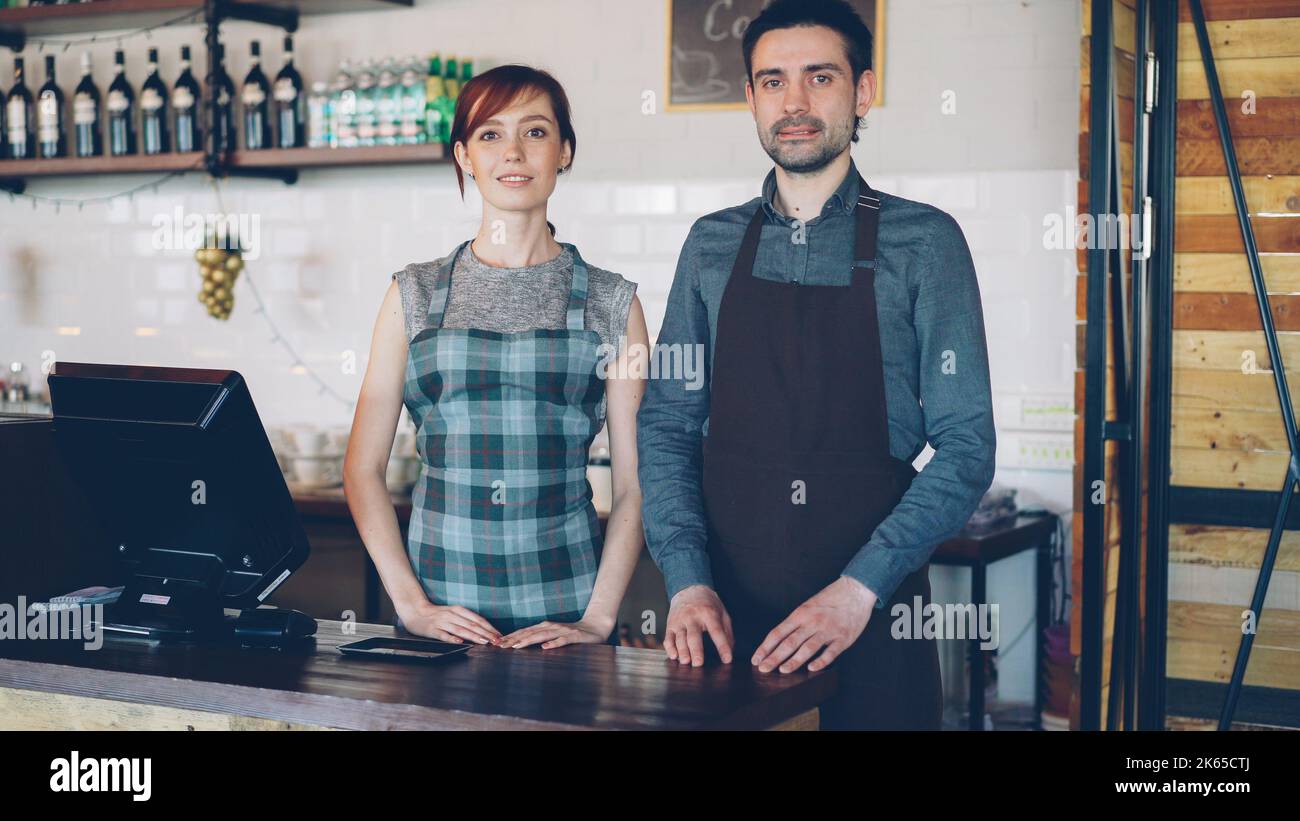 Portrait of two confident waiters in aprons standing at cashier's desk ...