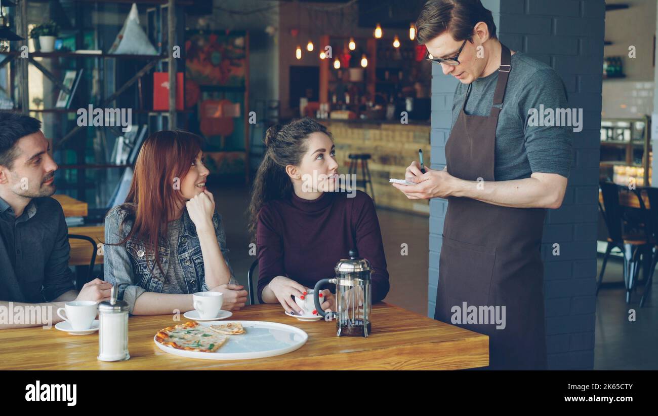 Friendly male waiter is taking order from cheerful group of friends ...