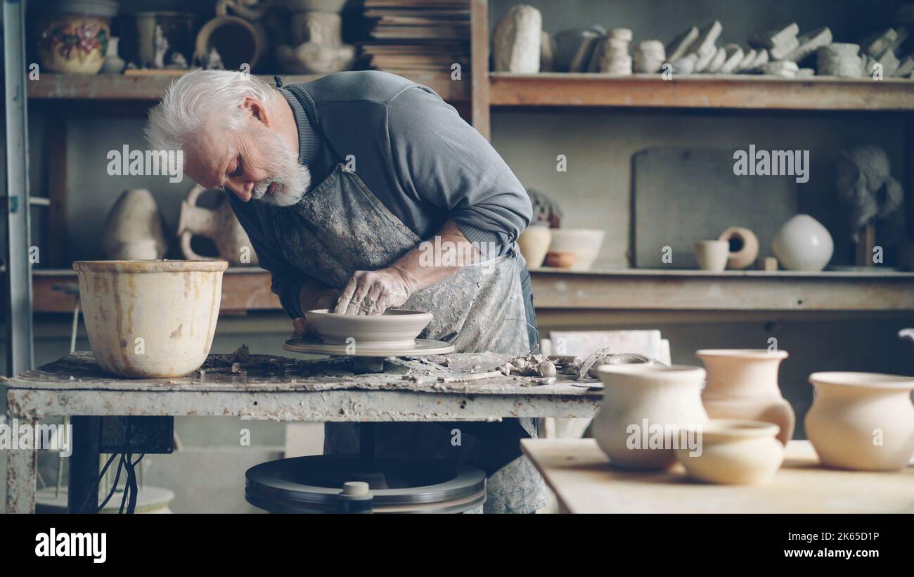 Experienced senior master is making low clay bowl on a pottery wheel in ...