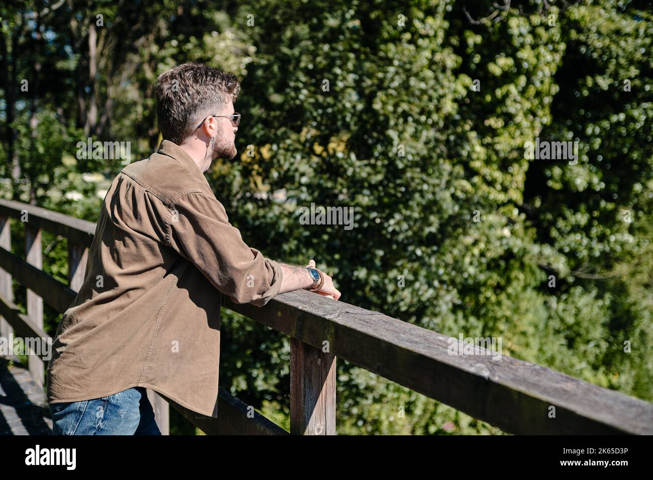 A blond guy leaning on the railing of a bridge in a forest looking ...