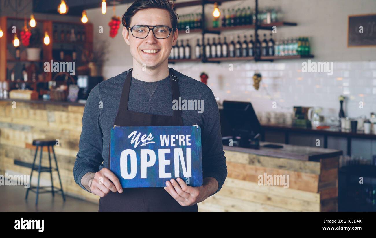 Portrait of cheerful handsome young waiter in apron holding we are open ...