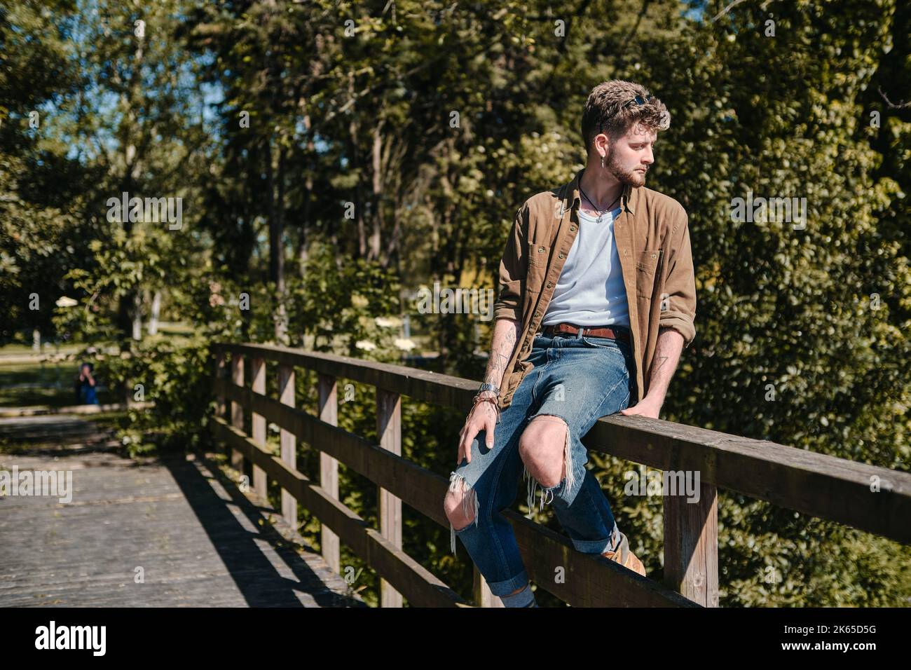 A blond guy sitting on the railing of a bridge looking aside with trees ...