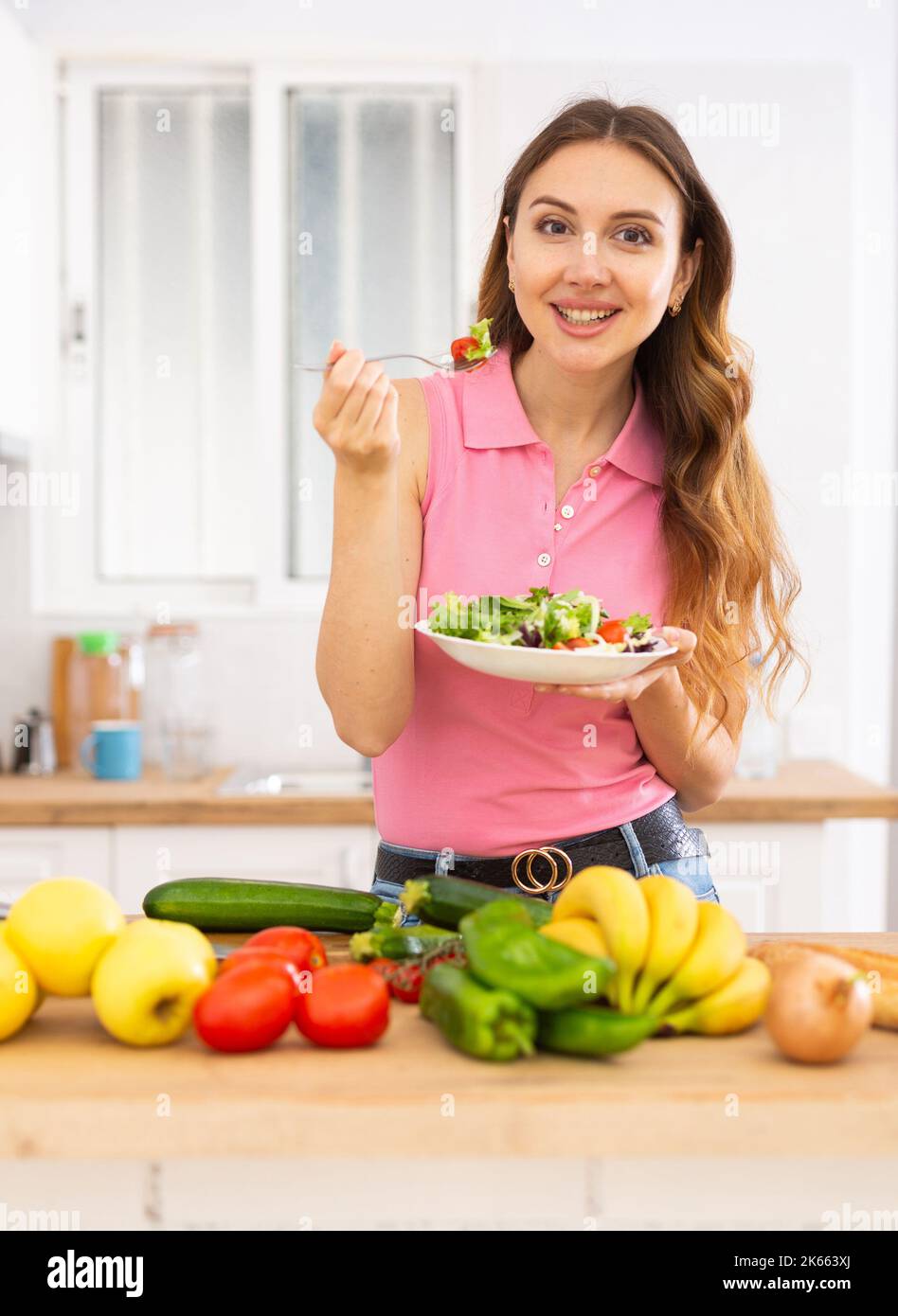 Vegeterian woman standing at table in kitchen and eating vegetable ...
