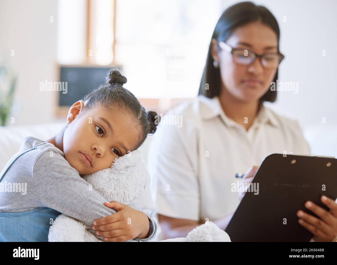 Sad little girl hugging her teddy bear while a social worker writes on ...