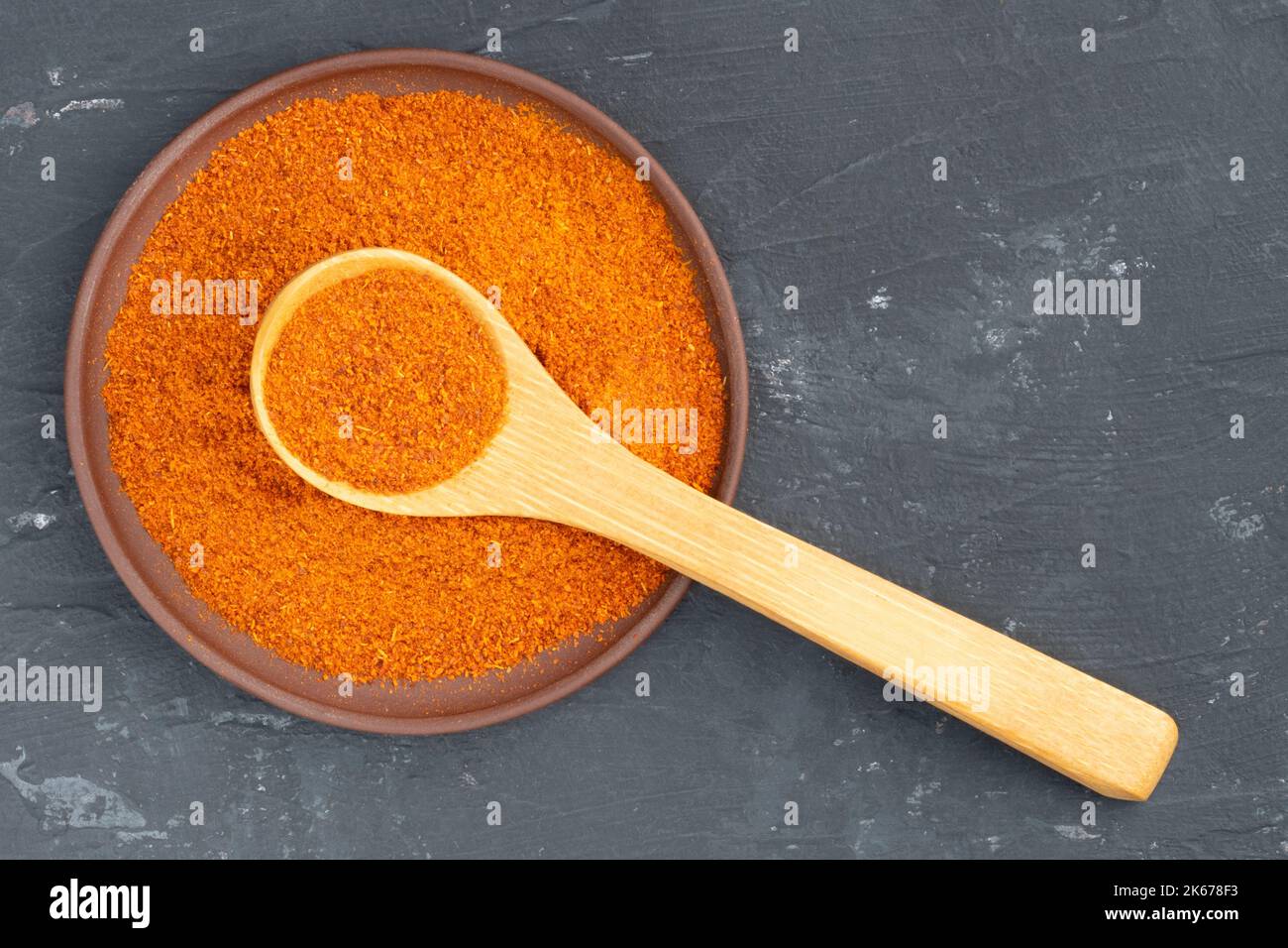 Red chili pepper spice in clay plate and wooden spoon on black concrete ...