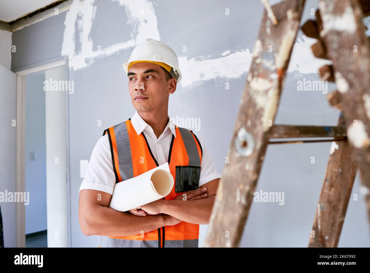 A serious construction worker stands with plans under his arm at home ...