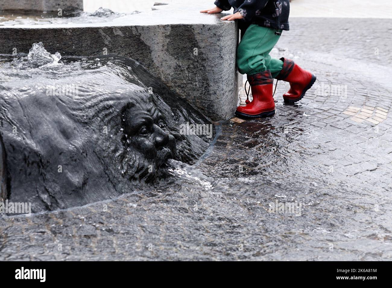 Fountain with water spitting sculpture and child in red rubber boots ...