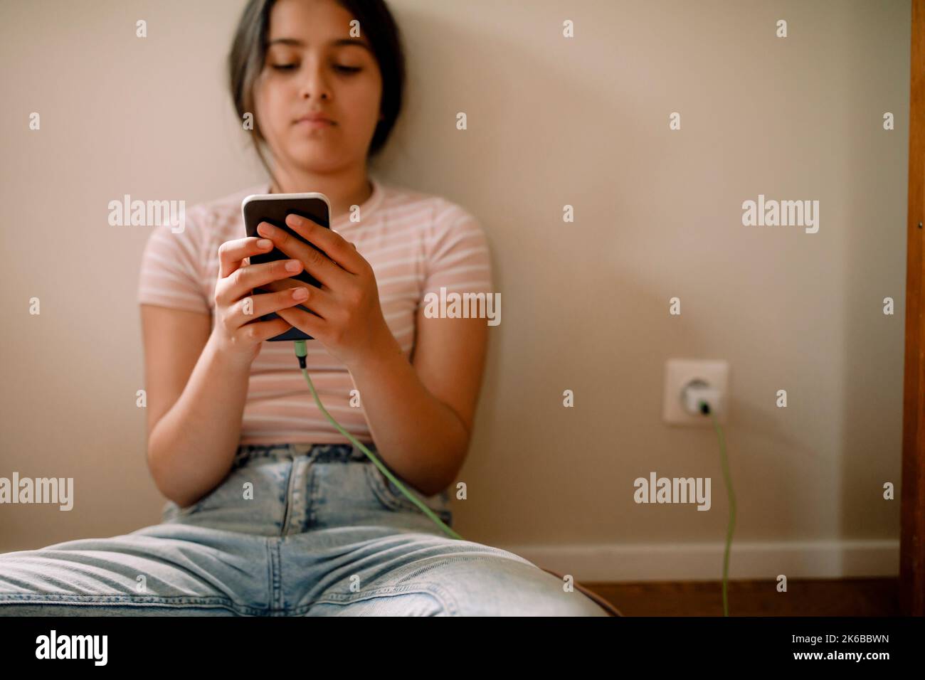 Girl charging and using mobile phone while sitting against wall at home ...