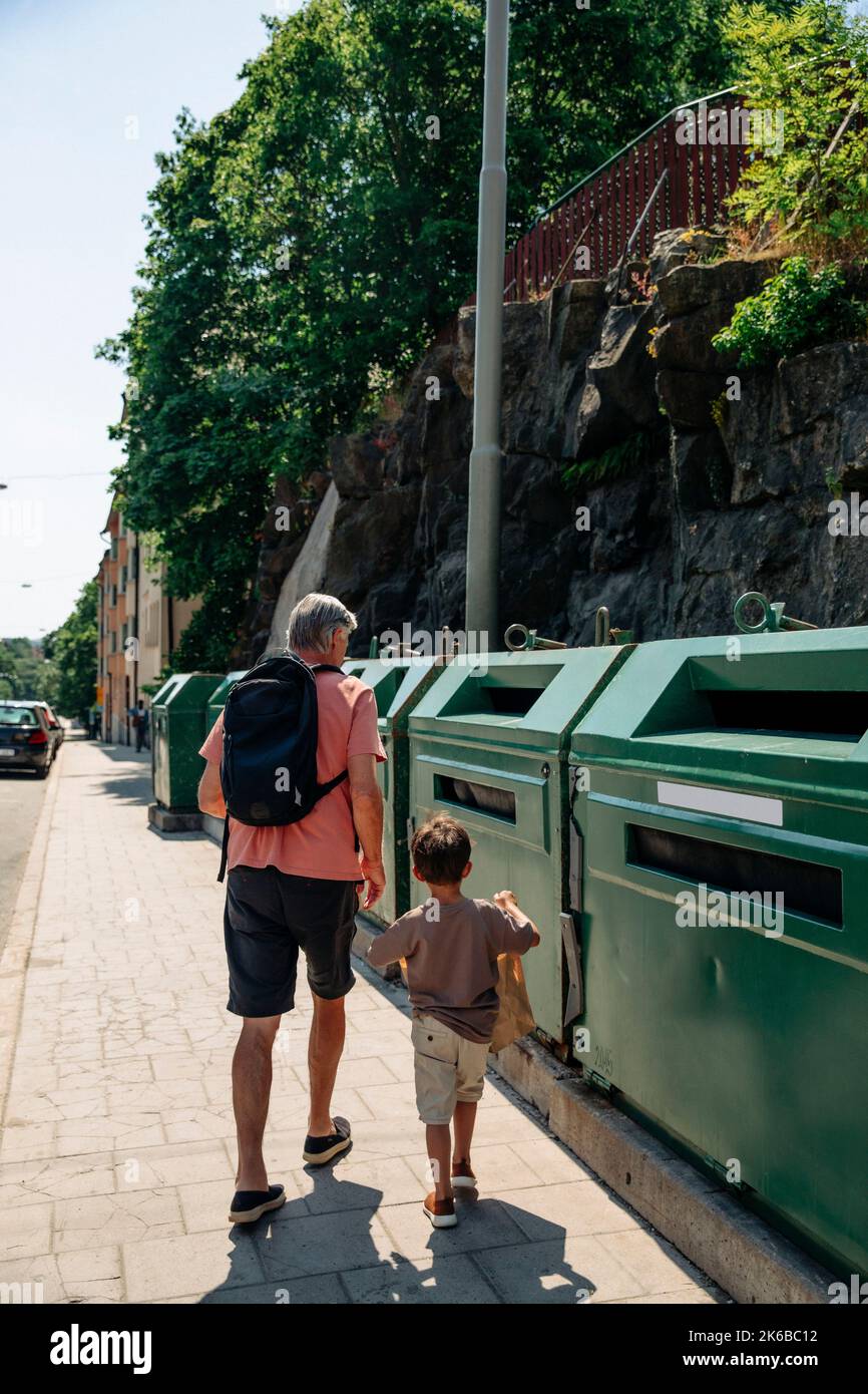 Rear view of grandfather and grandson walking towards garbage bins ...