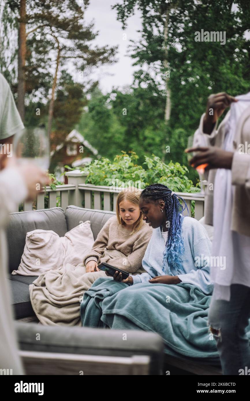 Girls with blanket sharing smart phone while sitting on couch during ...